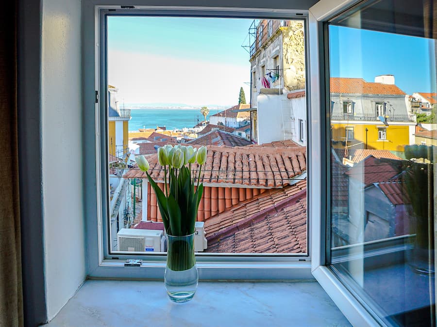 Janela com vista clara para uma vila costeira, telhado em azulejos e vaso com flores no parapeito.