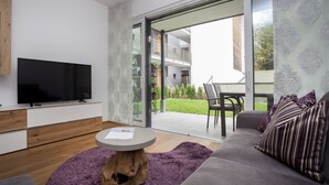 Uma sala de estar moderna com uma TV de tela plana, uma mesa de centro redonda com base de madeira, um sofá cinza e um tapete roxo.