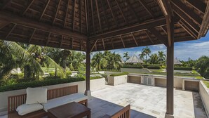 Uma área de estar coberta ao ar livre com móveis de madeira e vista para um jardim paisagístico com palmeiras.