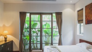 Quarto de hotel com cama, mesa de cabeceira com luminária, janela com cortinas e vista para o verde lá fora.