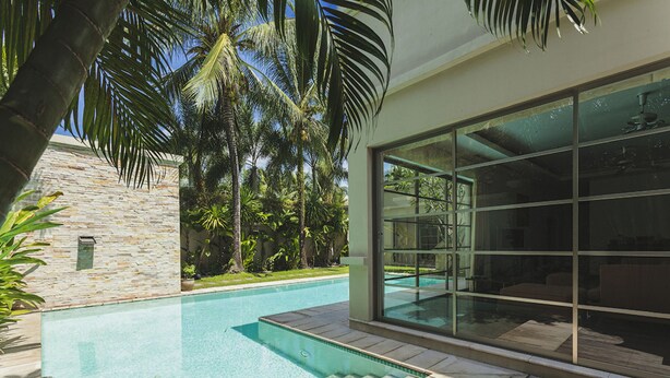 Uma área moderna de piscina externa com muro de pedra, palmeiras e um terraço com paredes de vidro.