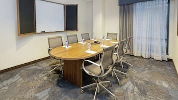 Uma sala de conferência com uma mesa redonda, seis cadeiras, um quadro branco e uma janela com cortinas.