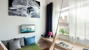Quarto moderno com sofá verde, rede, escrivaninha com luminária e uma grande janela com cortinas.
