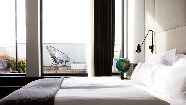 Quarto de hotel com uma cama grande, um globo, uma luminária de mesa e vista para uma varanda com cadeira e banco.