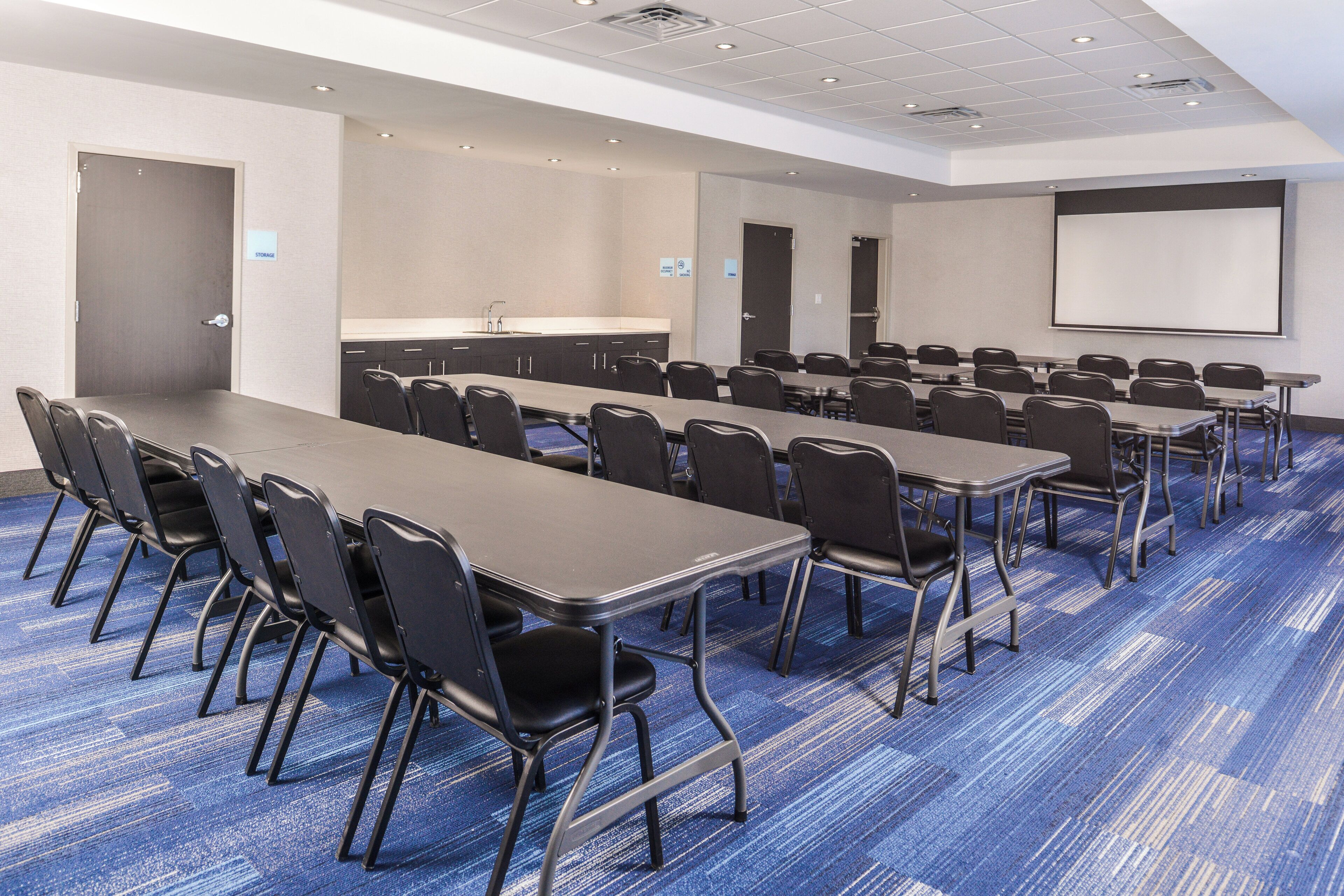 Sala de conferências com várias mesas e cadeiras, tela de projetor e um espaço com cozinha.