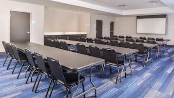 Sala de conferências com várias mesas e cadeiras, tela de projetor e um espaço com cozinha.