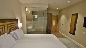 Quarto de hotel com uma cama grande, chuveiro em cabine de vidro, guarda-roupa de madeira e um tapete pequeno.
