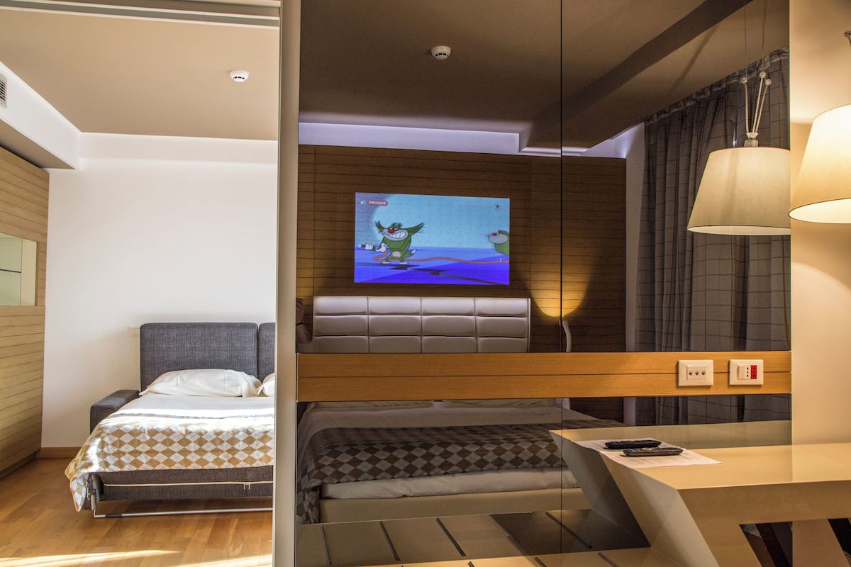 Quarto de hotel com uma cama, uma escrivaninha, televisão presa na parede e uma luminária.