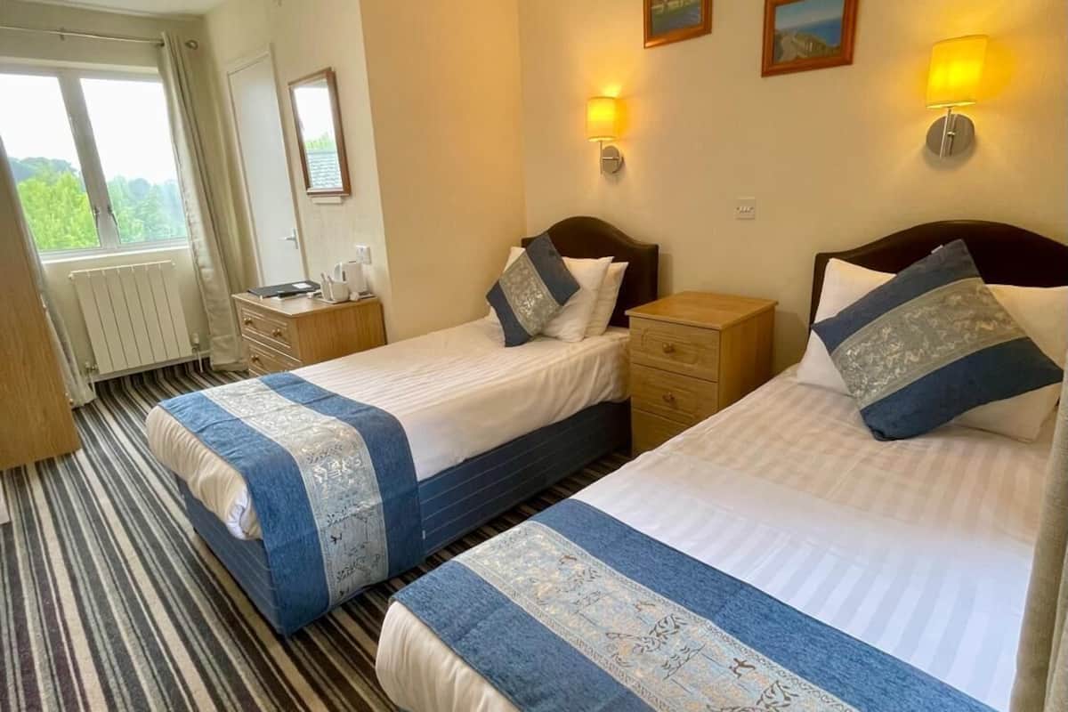 Quarto de hotel com duas camas, uma janela, um radiador, uma mesa de cabeceira de madeira e luminárias fixadas na parede.