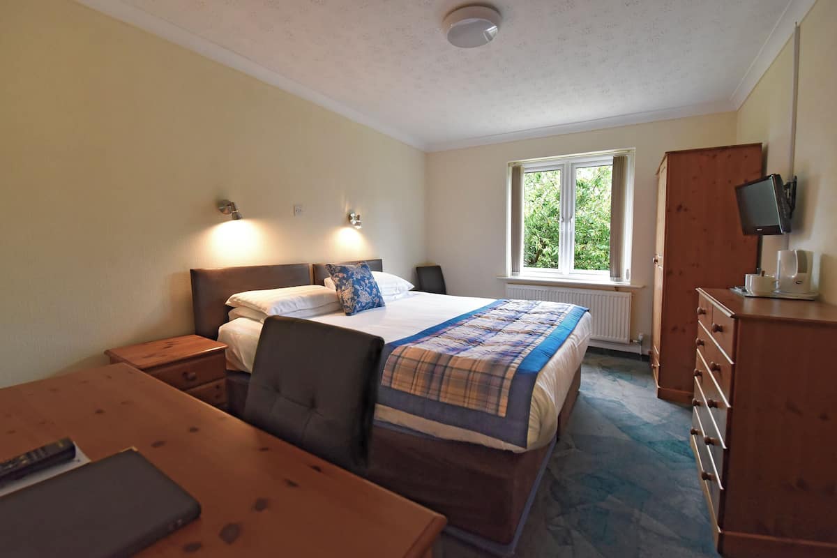 Quarto com cama, mesas de cabeceira, escrivaninha, cadeira, cômoda, televisão e janela com vista para área verde.