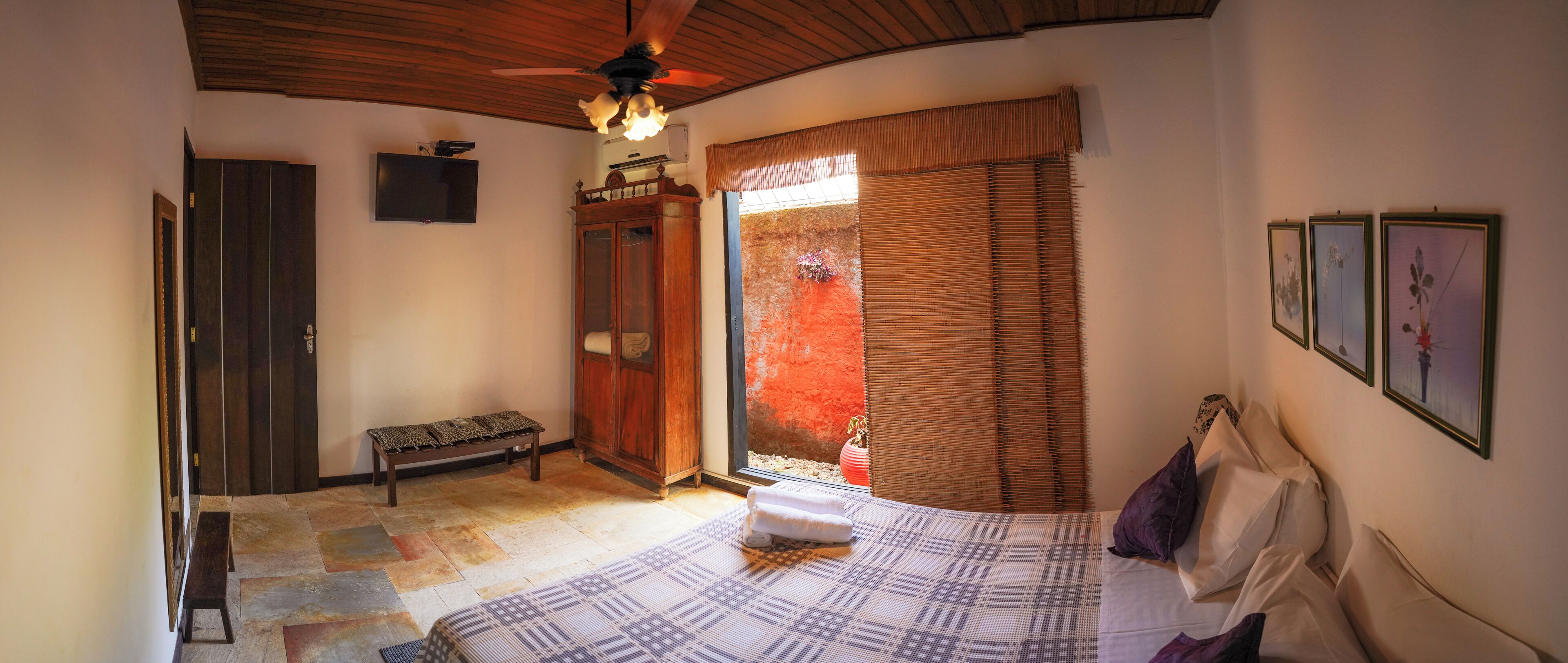 Quarto com cama, um banquinho, guarda-roupa, televisão e janela com persianas.