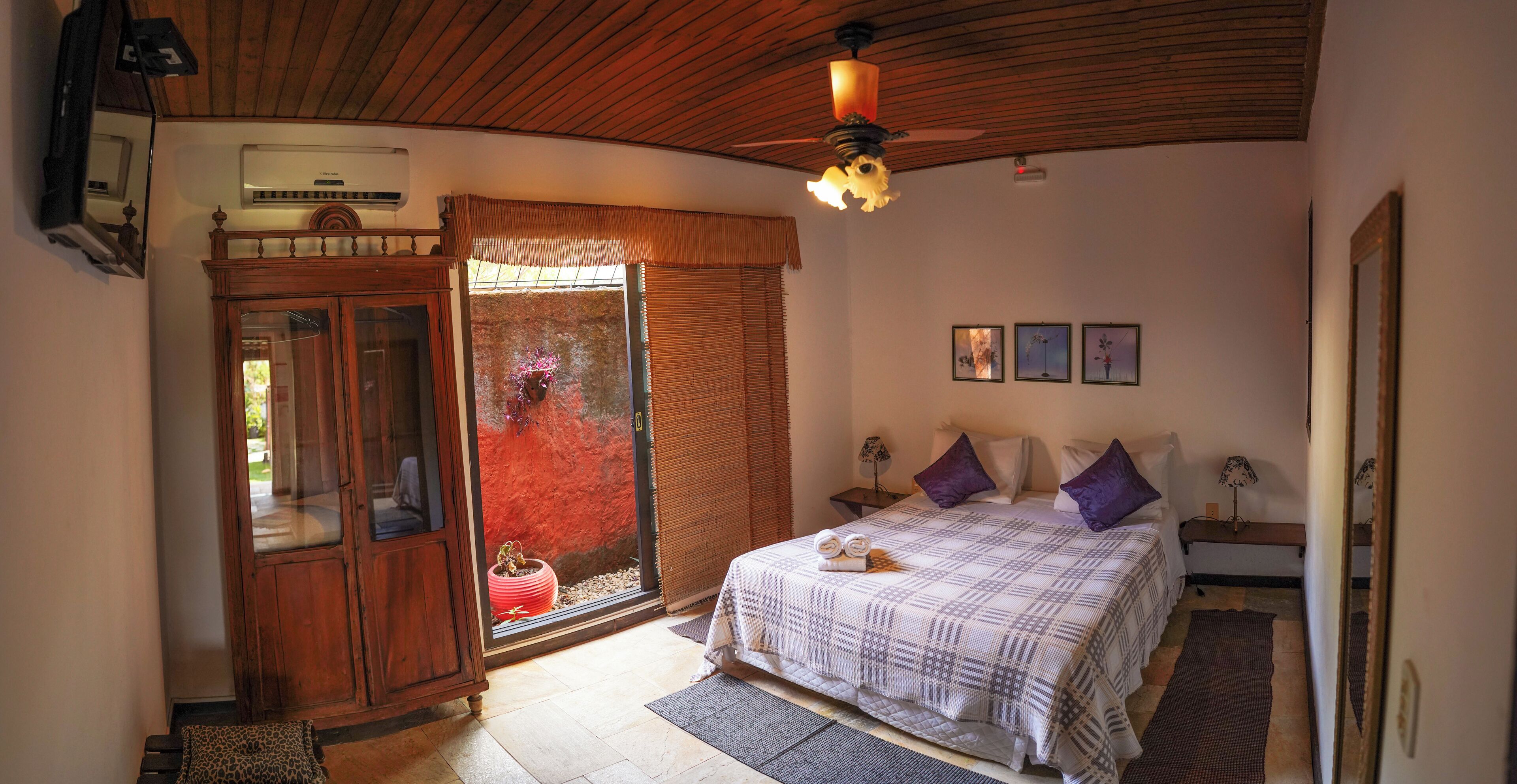 Quarto com cama, guarda-roupa de madeira, janela com vista e ventoinha no teto.