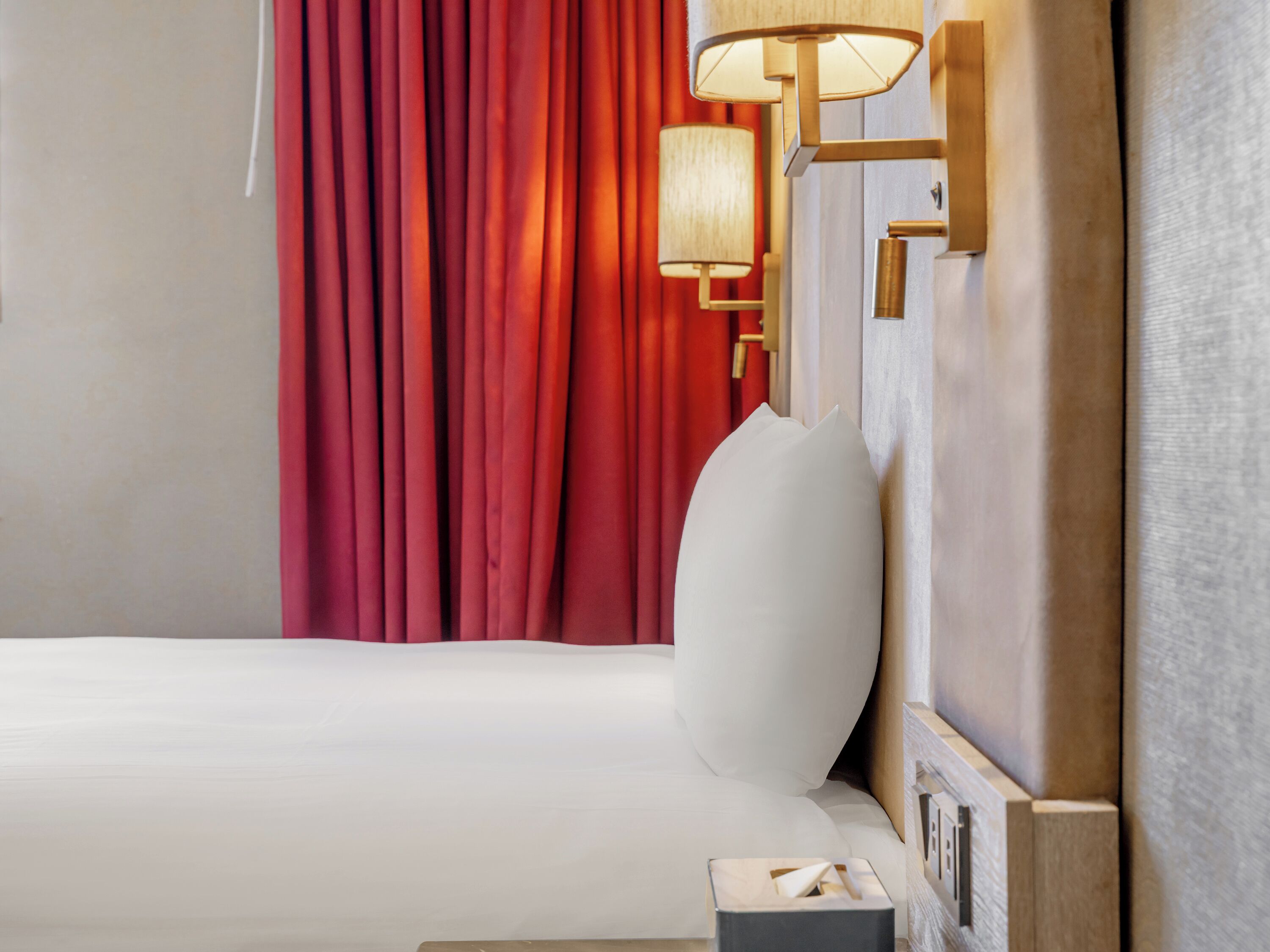 Quarto de hotel com cama, cortinas vermelhas, luminária na parede e mesa de cabeceira com caixa de lenços.