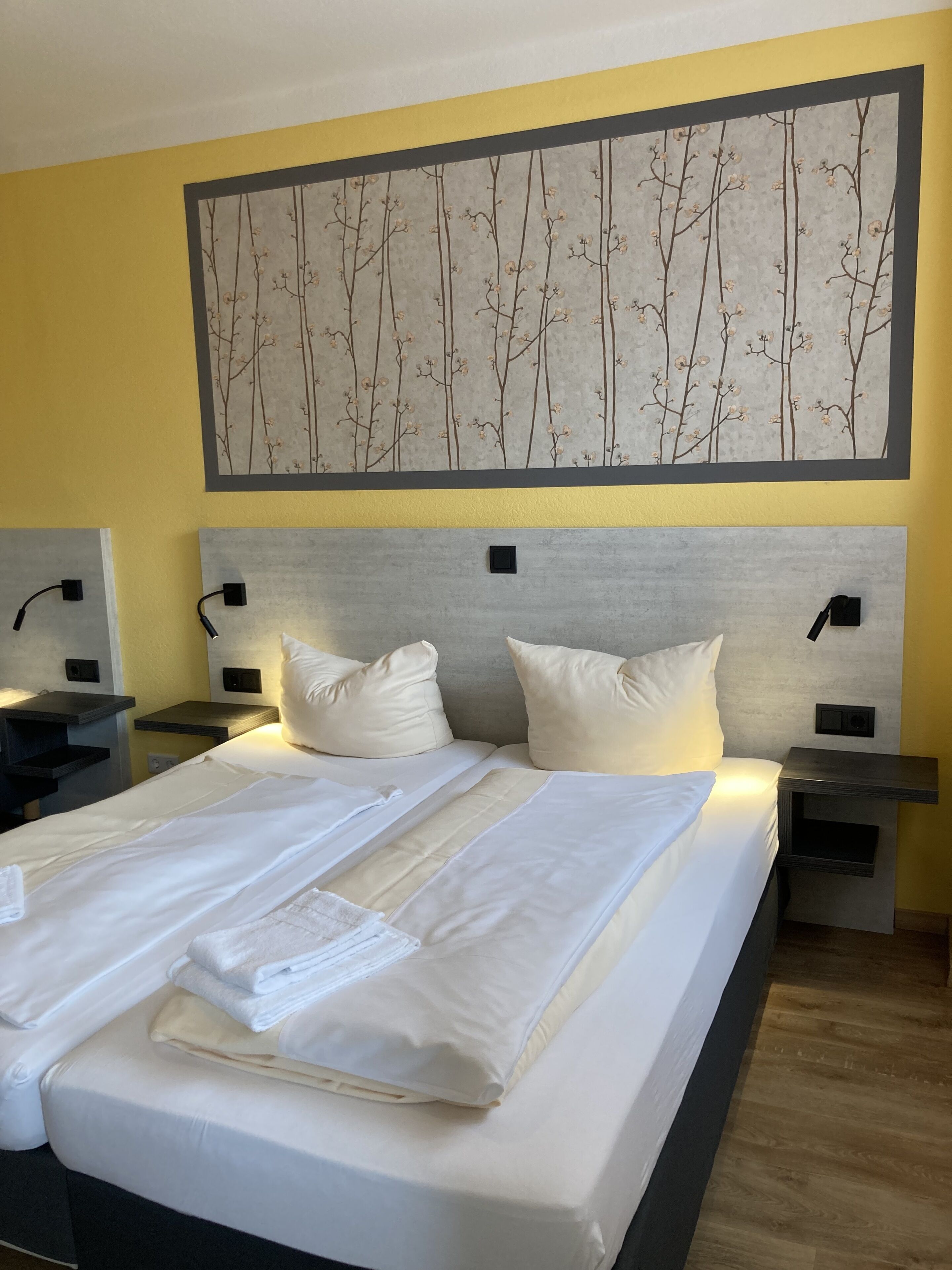 Cama bem arrumada com lençóis e almofadas brancas, uma grande obra de arte na parede com galhos e flores, e mesas de cabeceira com luminárias.