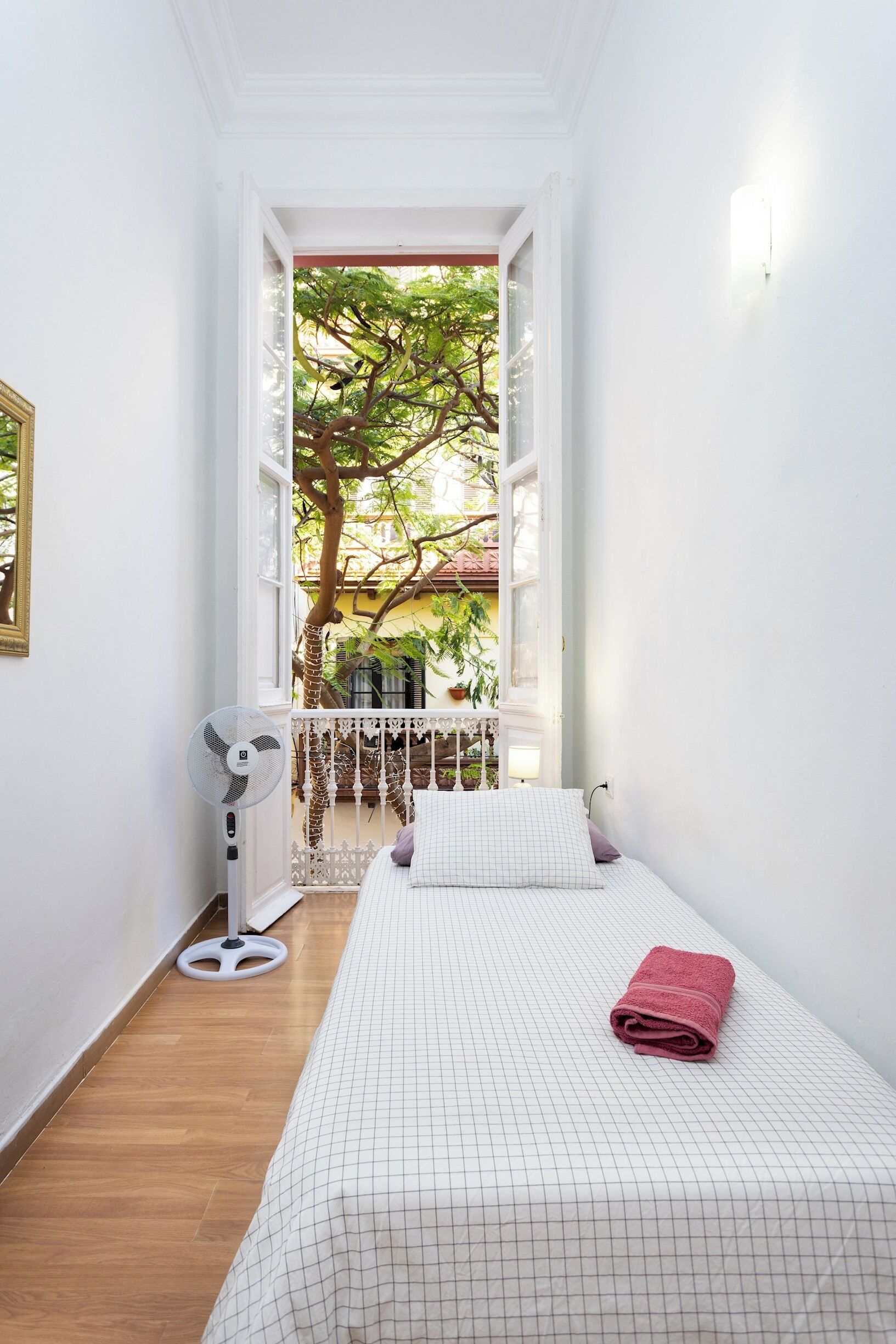 Um quarto pequeno com uma cama, um ventilador e uma janela que dá para uma varanda com uma árvore.