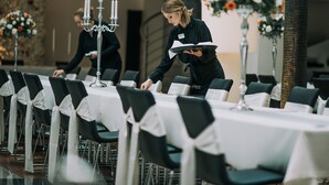 Uma sala de banquetes com mesas dispostas em fileiras, cada uma com toalha branca e cadeiras pretas.