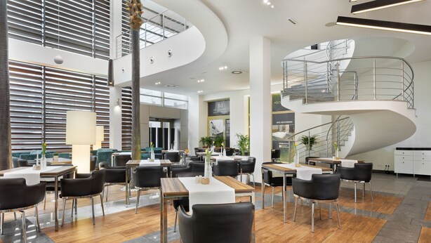 Um amplo hall de hotel com design moderno, apresentando uma área central de estar com mesas e cadeiras, uma escada em espiral e janelas grandes com persianas.