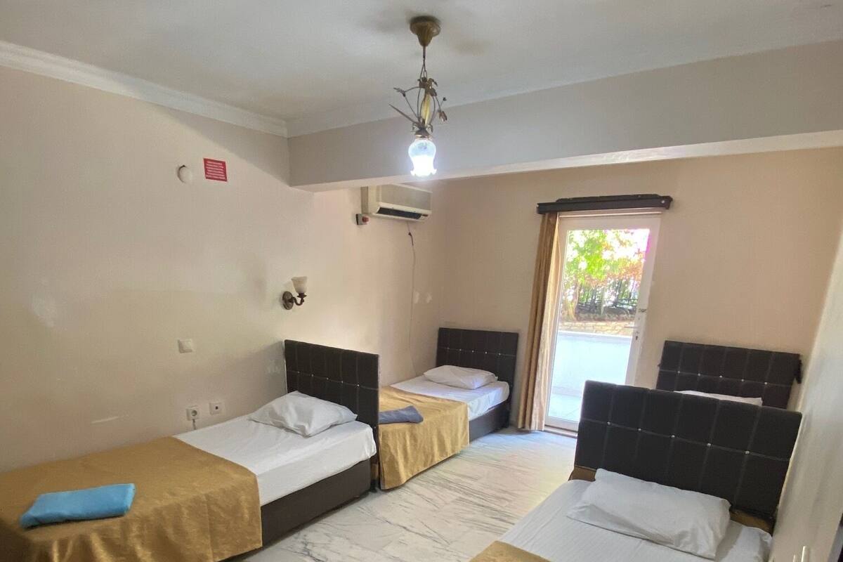Quarto de hotel com três camas de solteiro, um ventilador de teto, ar condicionado e vista para a rua através de uma porta.