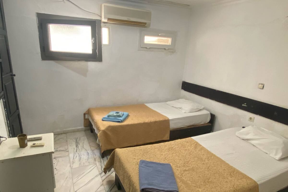 Quarto pequeno e simples com duas camas de solteiro, uma escrivaninha e uma janela.
