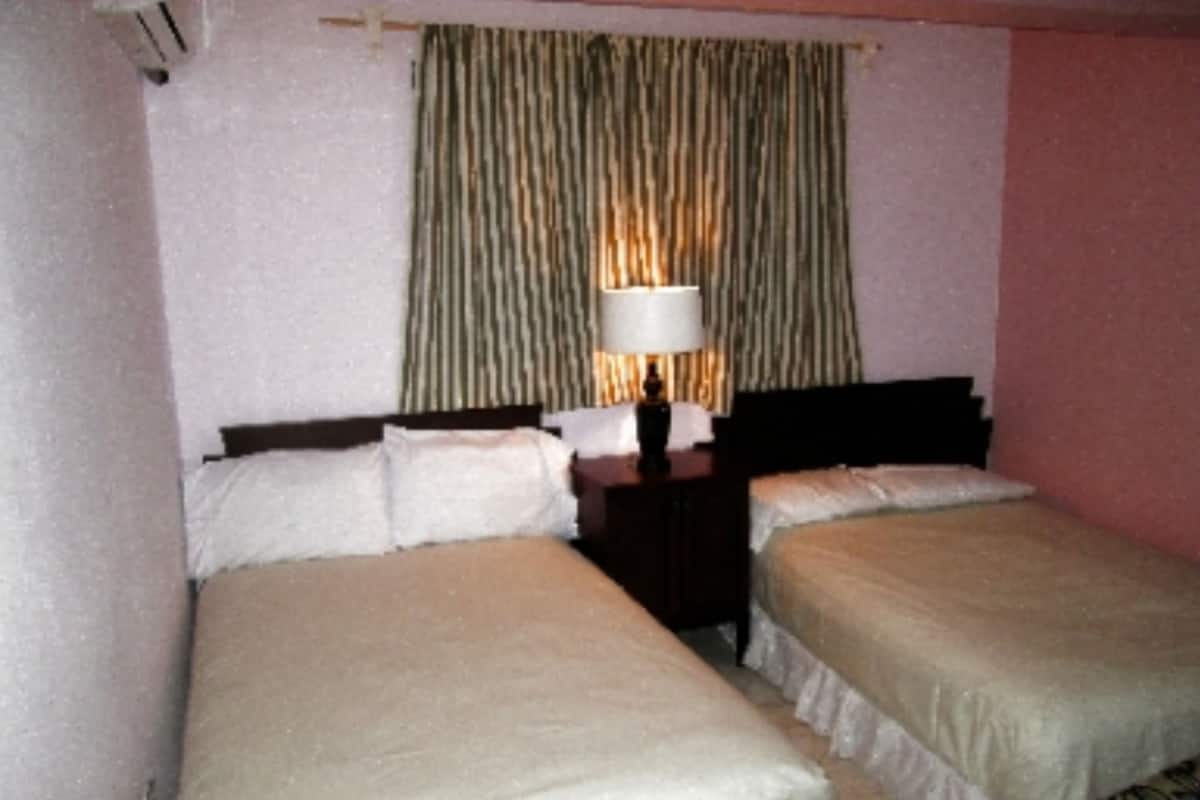 Quarto de hotel com duas camas de solteiro, uma mesa de cabeceira com abajur e uma janela com cortinas listradas.