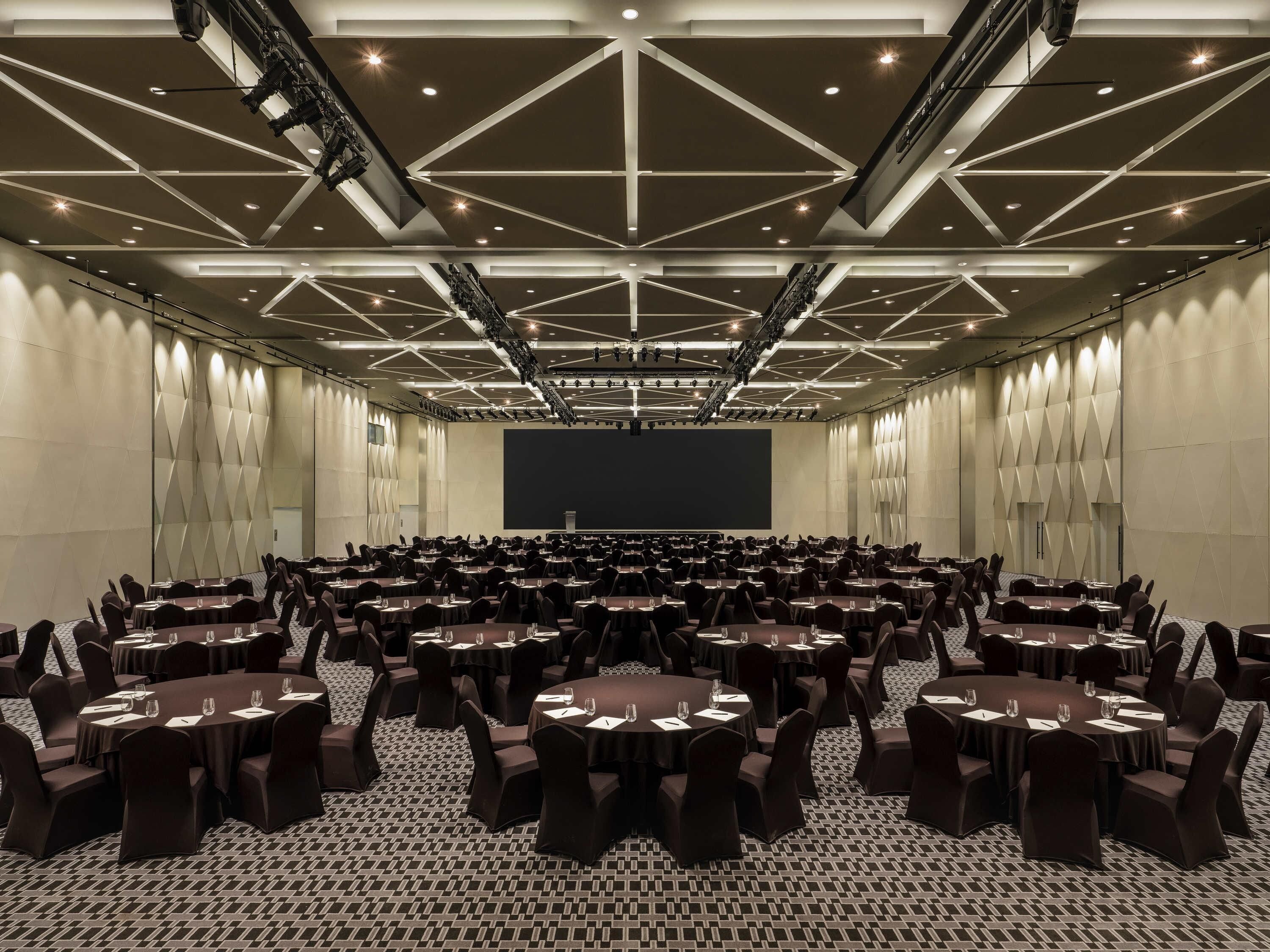 Uma sala de conferência com várias mesas redondas, cadeiras, uma tela grande e um palco.