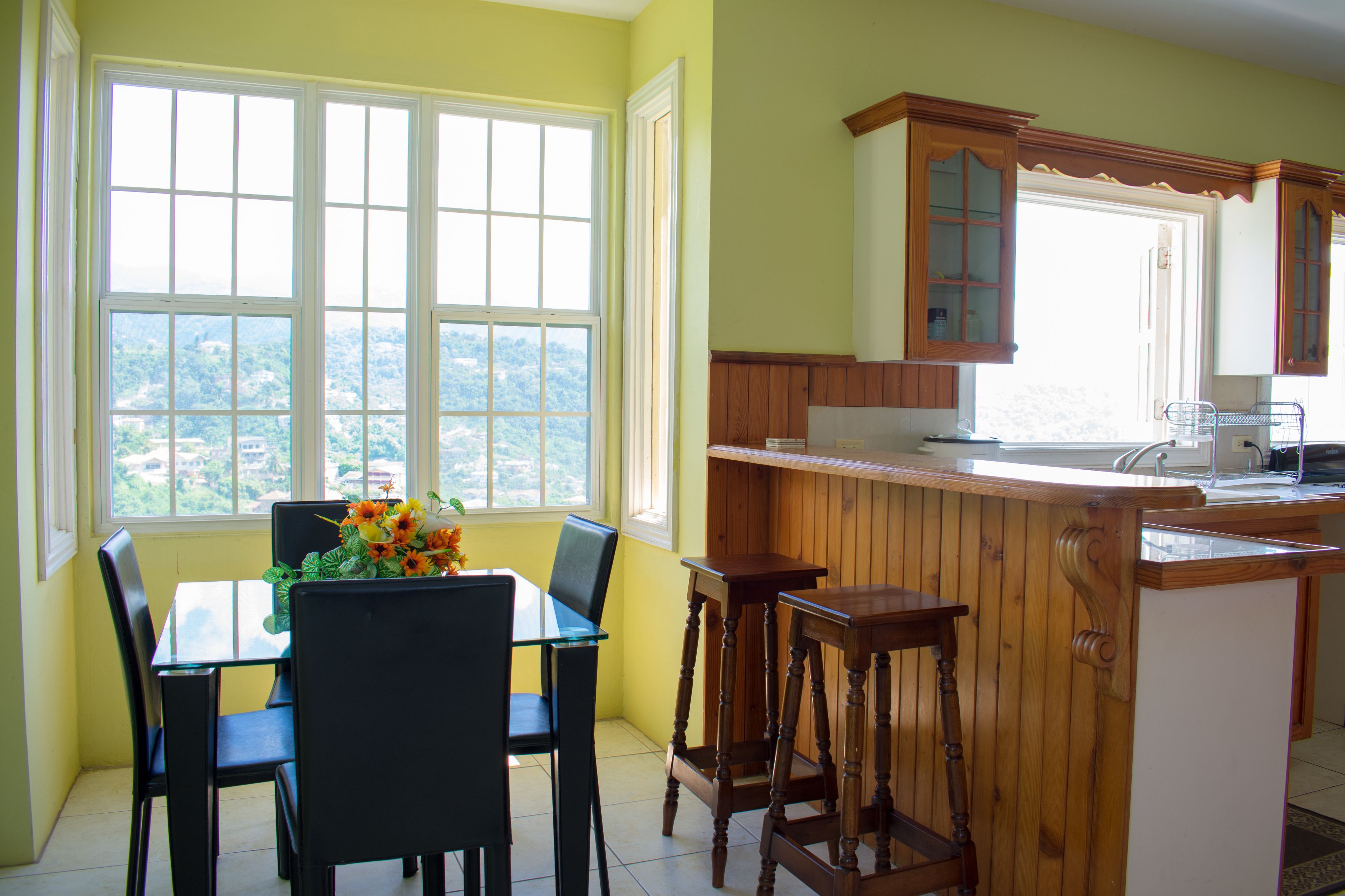 Uma área de refeições com mesa e cadeiras, uma cozinha com armários de madeira e uma janela com vista para o verde.