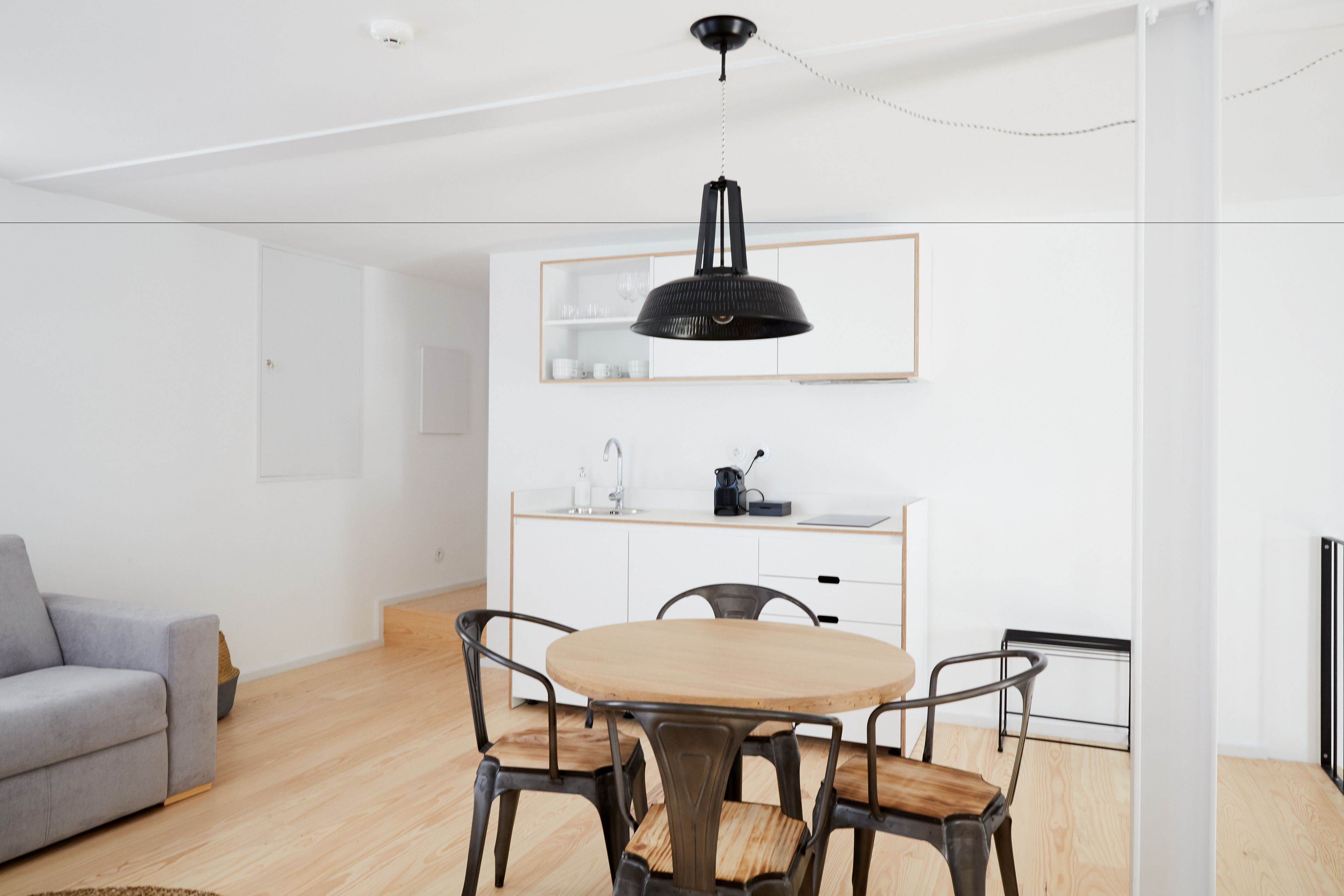 Uma sala de estar moderna com uma mesa de madeira redonda, cadeiras de metal, um sofá cinzento e um aparador branco com uma máquina de café.