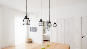 Quarto moderno com mesa de jantar de madeira, três luminárias pendentes e vista para outro quarto com uma cama.
