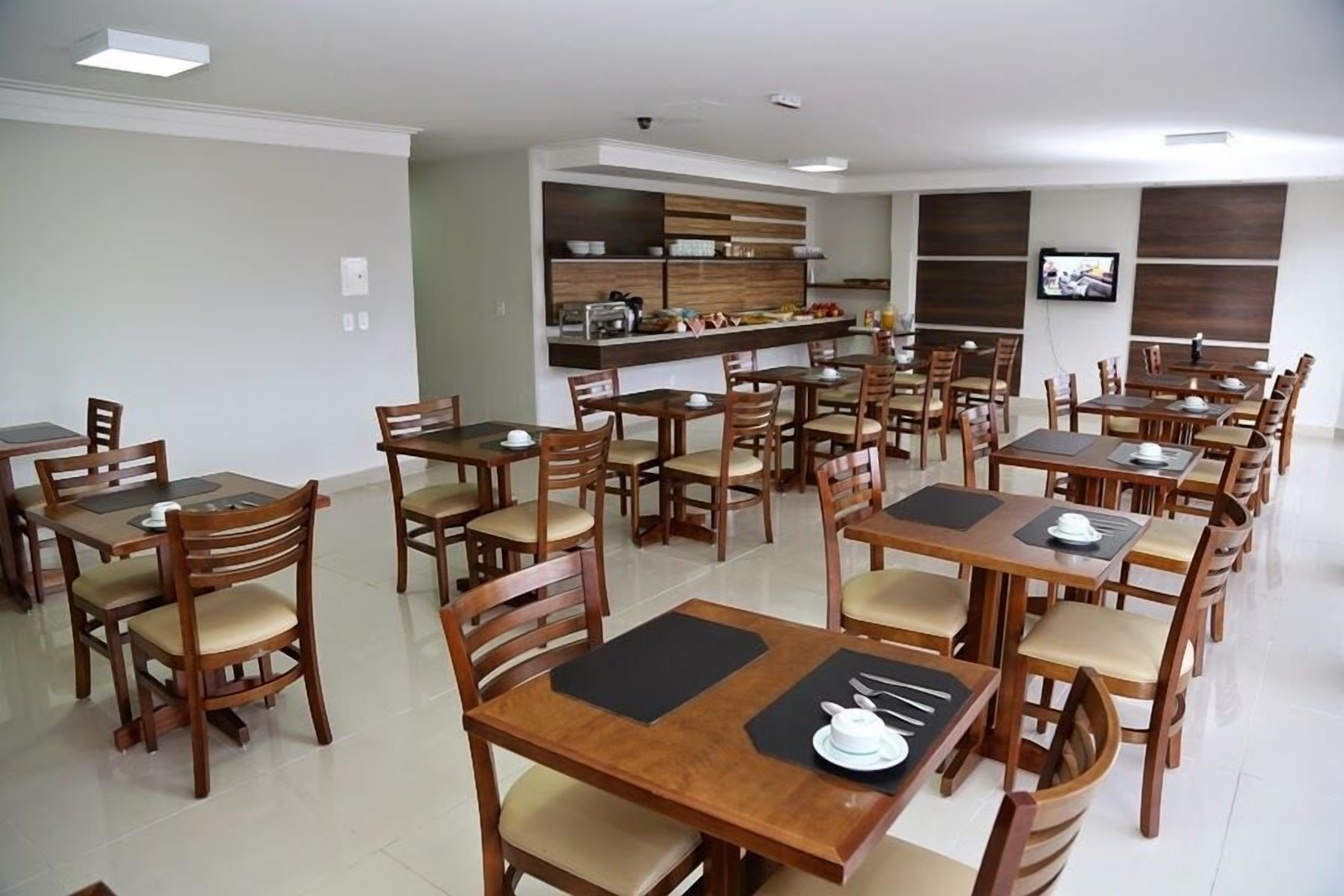 Uma sala de jantar com várias mesas e cadeiras, uma cozinha com eletrodomésticos e prateleiras, e uma televisão fixada na parede.