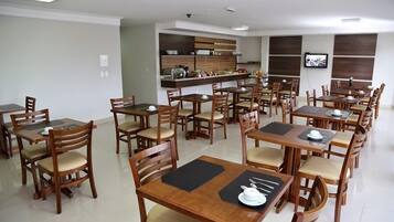 Uma sala de jantar com várias mesas e cadeiras, uma cozinha com eletrodomésticos e prateleiras, e uma televisão fixada na parede.