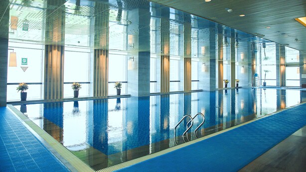 Piscina interior com paredes de vidro, plantas em vasos e borda azul.