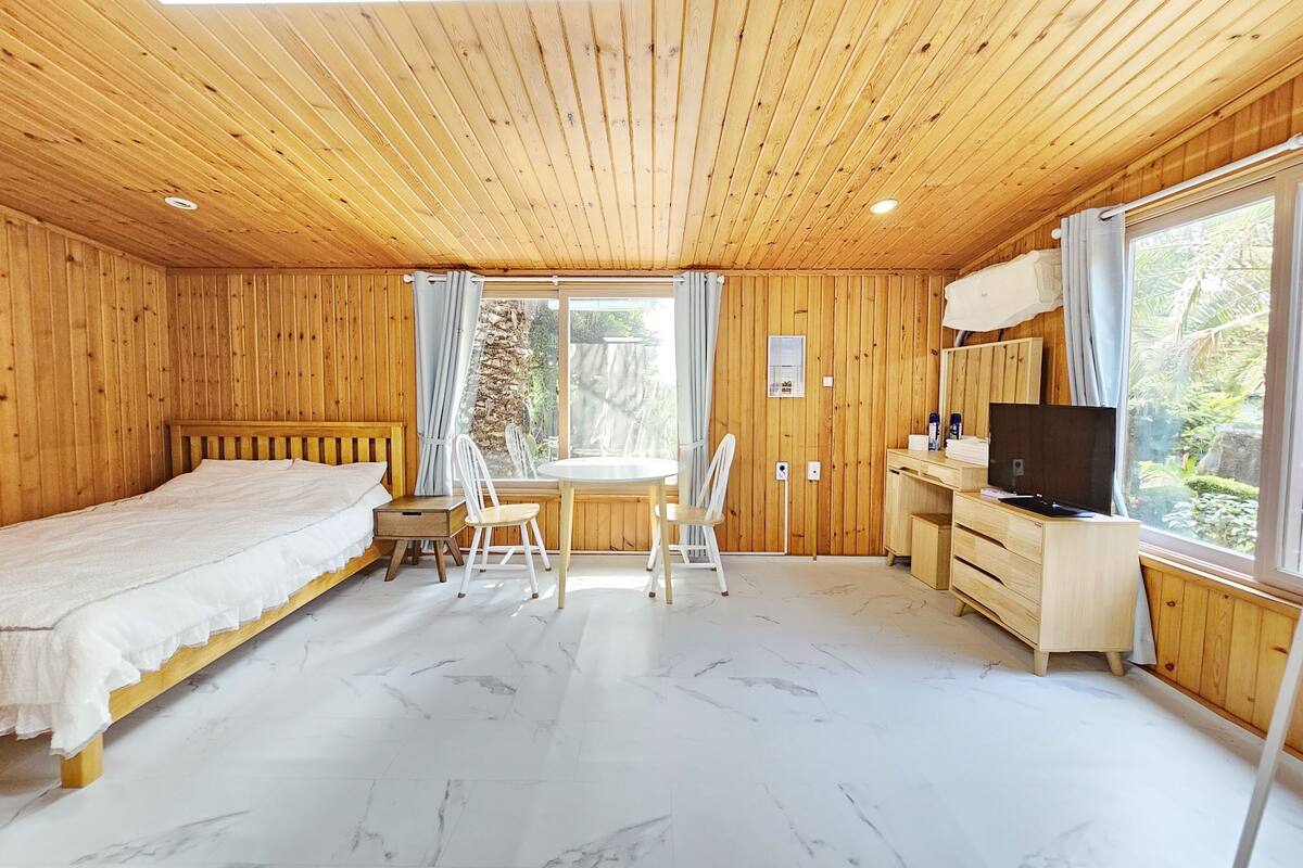 Quarto de cabana de madeira com uma cama, uma escrivaninha com cadeira, uma televisão e janelas grandes.