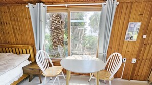 Quarto acolhedor com uma cama, uma mesa pequena, duas cadeiras brancas e uma janela com vista para uma árvore e vegetação.