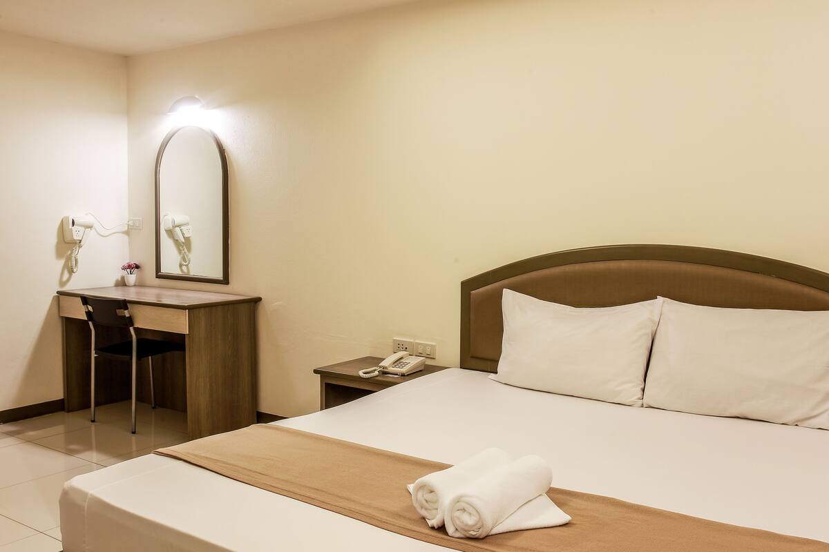 Quarto de hotel com cama, escrivaninha, cadeira, espelho e mesa de cabeceira com telefone.