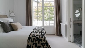 Um quarto com cama, janela com cortinas, radiador e banheiro visível por uma porta aberta.