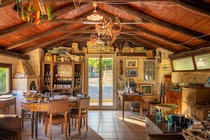 Um interior rústico com teto de madeira, área de jantar com mesa e cadeiras, estante cheia de livros e garrafas, e um pequeno altar com objetos religiosos.