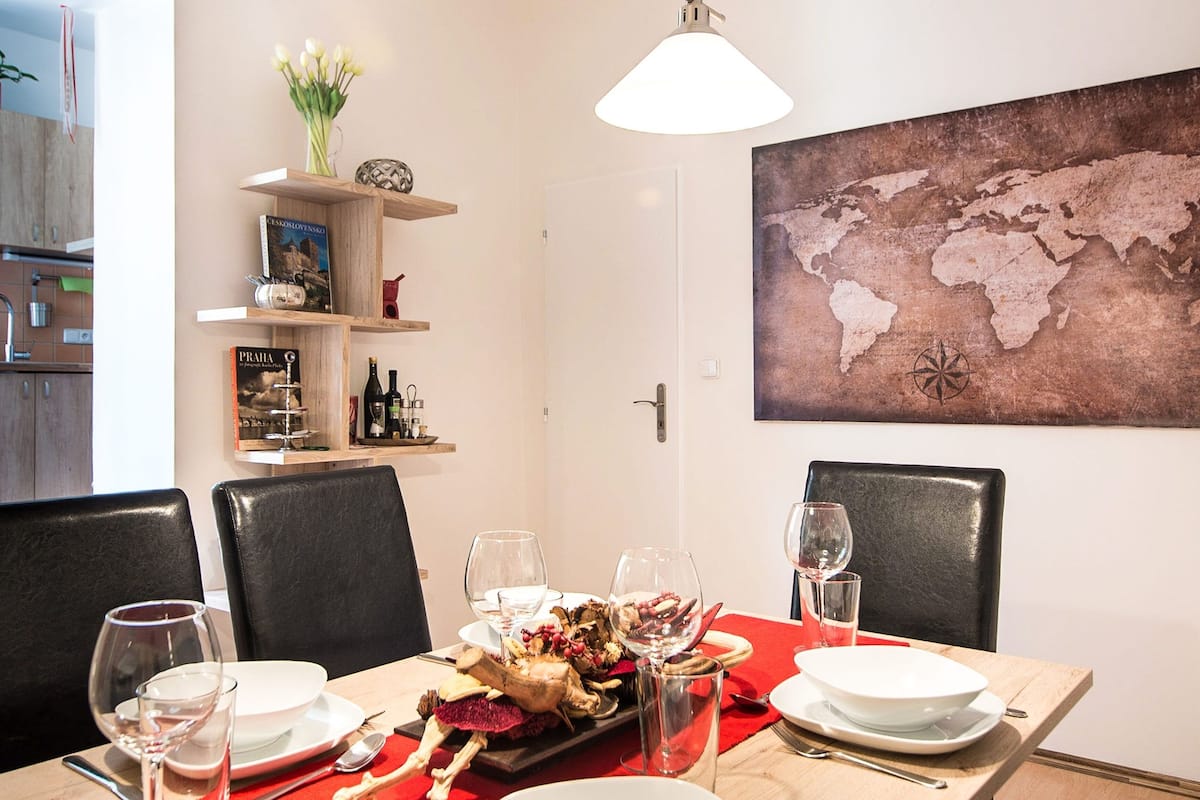 Uma sala de jantar com mesa de madeira posta para dois, um mapa-múndi na parede e uma cozinha ao fundo.