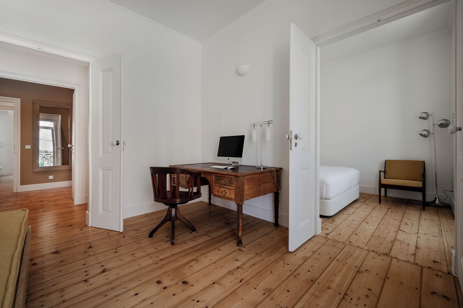 Quarto com escrivaninha de madeira, cadeira e cama. Há uma porta que dá para outro quarto.