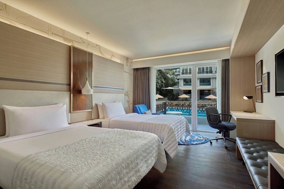 Quarto de hotel com duas camas, uma escrivaninha, uma cadeira, um sofá e vista para uma piscina e um edifício.