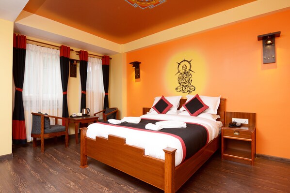 Quarto de hotel bem arrumado com uma cama de madeira, uma mesinha com chaleira, uma cadeira e uma mesinha de cabeceira com abajur.