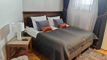 Quarto bem arrumado com cama, mesa de cabeceira, abajur e vista para o exterior através de cortinas leves.