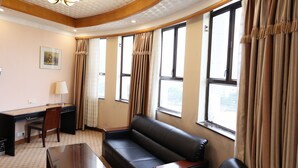 Quarto de hotel com escrivaninha, cadeira, sofá e janelas amplas com cortinas.