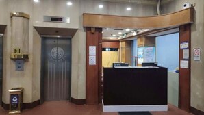 Uma área de recepção de hotel, com balcão, relógio digital e elevador.