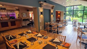 Restaurante com mesas e cadeiras de madeira, um bar e amplas janelas com vista para o exterior.