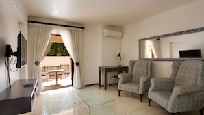 Uma sala de estar moderna com uma mesa de centro de vidro, duas poltronas estampadas, uma televisão e uma escrivaninha.