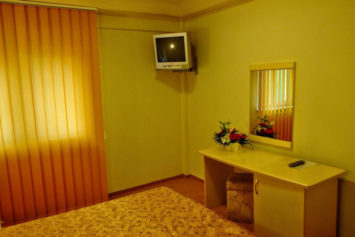 Quarto de hotel com cama, escrivaninha, televisão e espelho.
