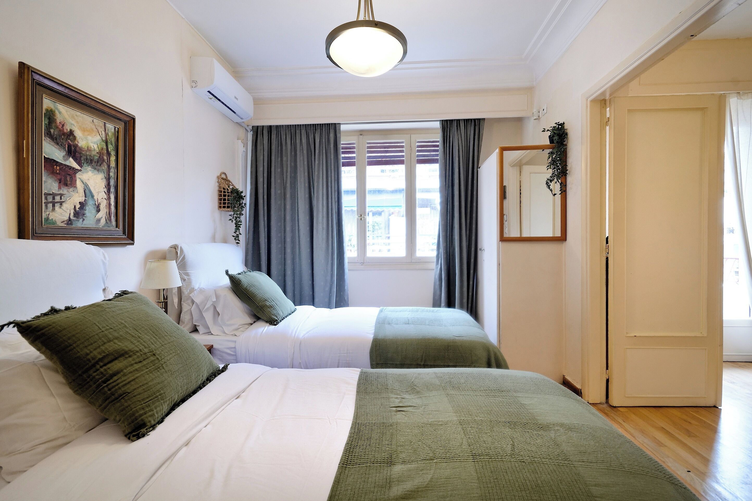 Quarto de hotel com duas camas, uma janela grande com cortinas, um quadro na parede e um ar condicionado.