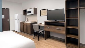 Quarto de hotel com cama, cozinha compacta com micro-ondas, televisão, escrivaninha e guarda-roupa.