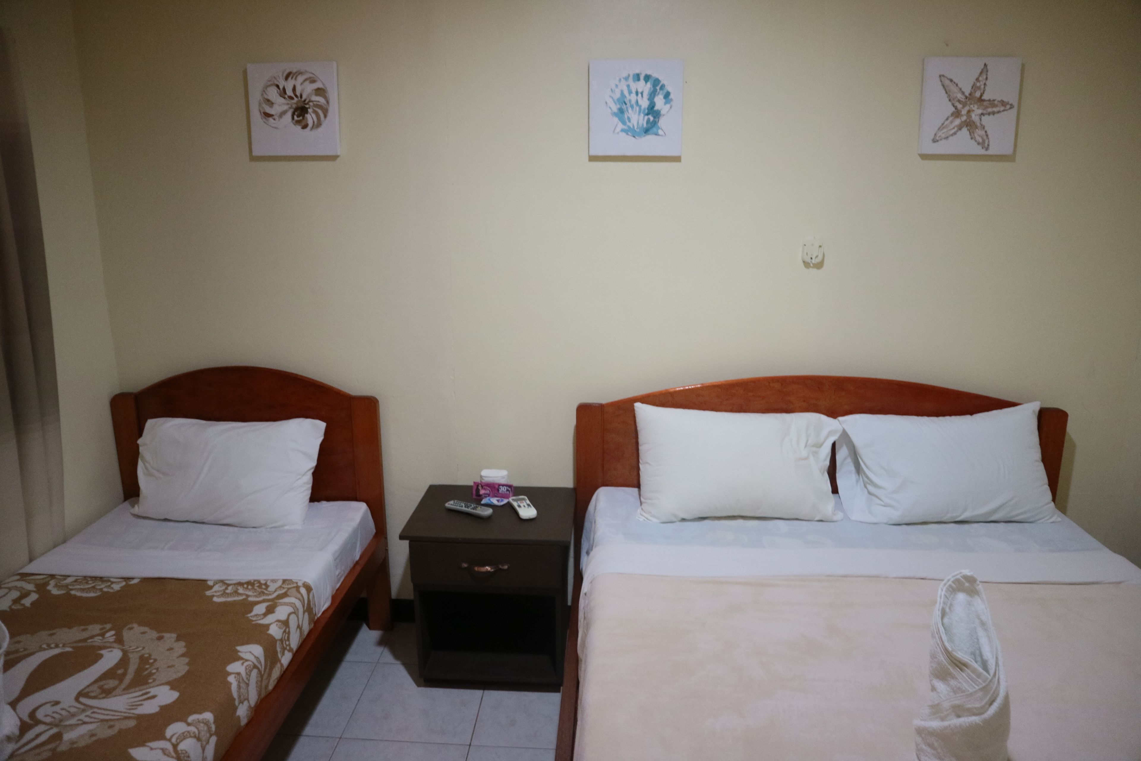Quarto de hotel com duas camas de solteiro, uma mesinha de cabeceira com controle remoto e obras de arte nas paredes.