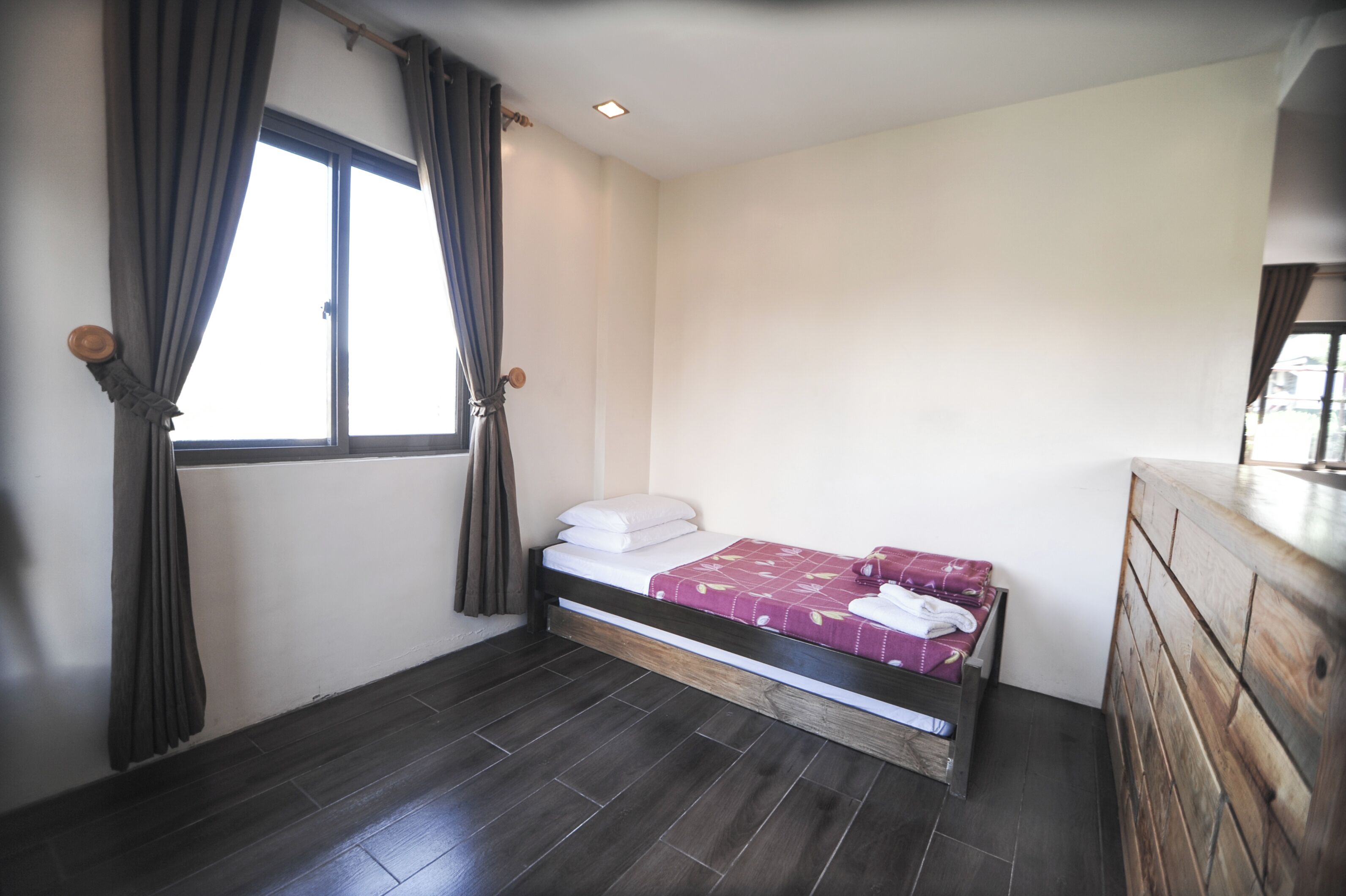 Quarto pequeno e bem arrumado com uma cama de solteiro, cômoda de madeira e janela com cortinas.